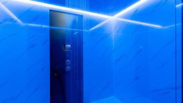 Um elevador com painel de controle e porta de vidro, inserido em um espaço com iluminação azul e paredes de mármore.