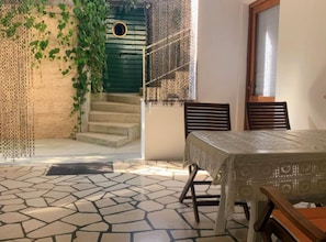 Um pátio com piso de pedra, uma mesa com toalha, duas cadeiras, uma escadaria e uma porta verde com janela circular.