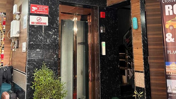 Entrada de hotel com elevador de vidro, placa de 'Proibido Fumar', vigilância por CCTV e placa de 'Sanitários'.