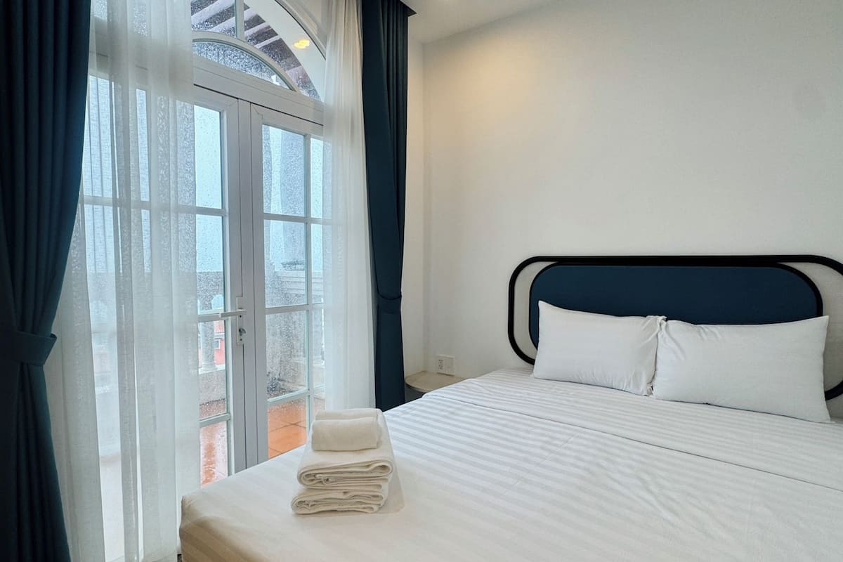 Quarto de hotel com cama, janela com cortinas, uma mesinha e vista para o exterior.