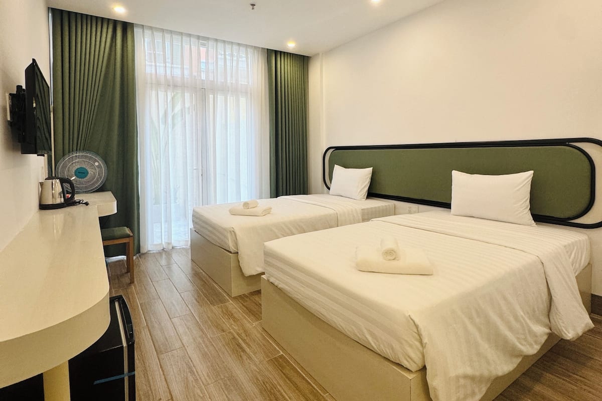 Quarto de hotel com duas camas, uma mesinha, um ventilador e uma televisão na parede.