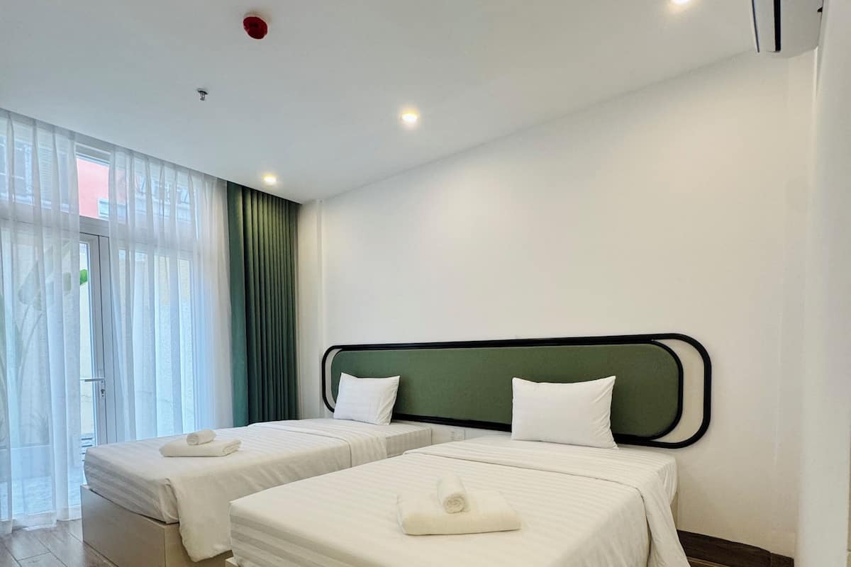 Quarto de hotel com duas camas, uma janela grande com cortinas leves, piso de madeira e teto com iluminação embutida.