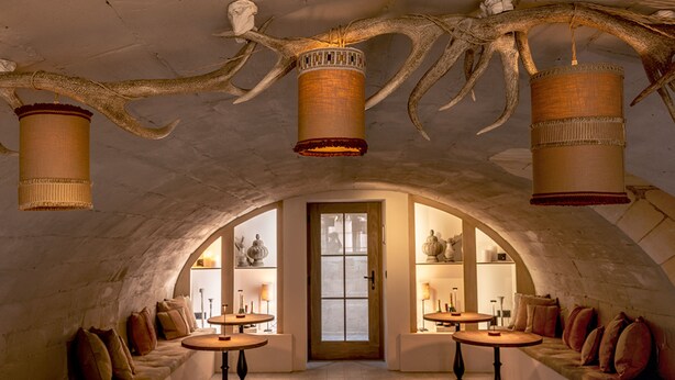 Teto abobadado com chifres de veado e luminárias pendentes, porta de madeira e um espaço de estar com mesas e almofadas.