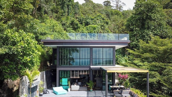Uma casa moderna de dois andares, com grandes janelas, um pátio com móveis de exterior e vegetação exuberante ao redor.