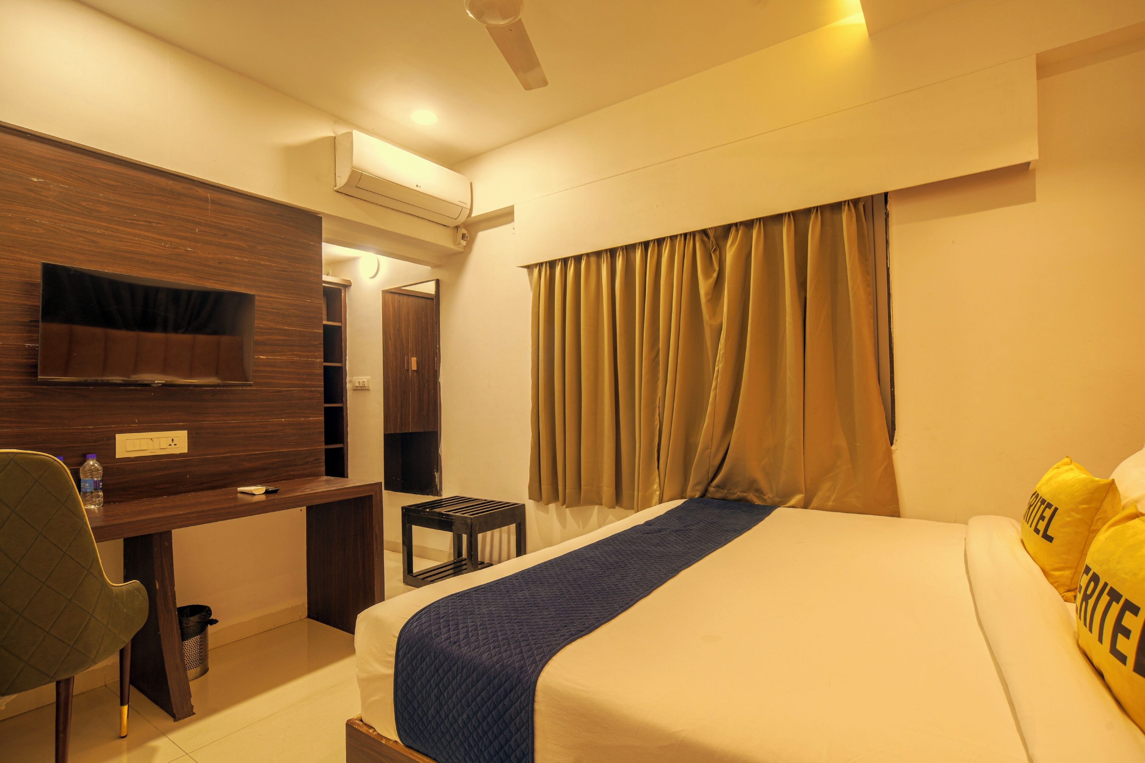 Quarto de hotel com cama, escrivaninha, cadeira, televisão e janela com cortinas.