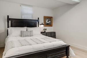 Quarto com cama de madeira escura, roupa de cama branca, abajur, quadro na parede e janela com persianas.