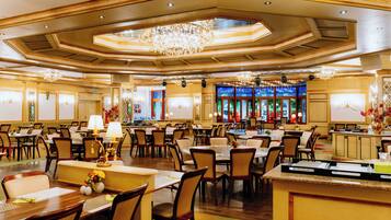 Ampla sala de jantar de hotel, com várias mesas, cadeiras, lustres e um bar.