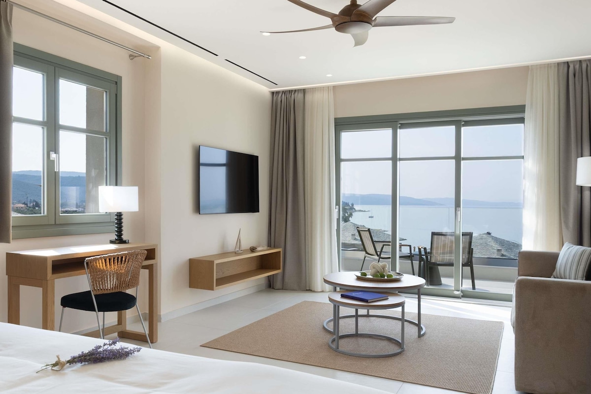 Quarto de hotel com cama, escrivaninha, televisão, vista para o mar e ventoinha no teto.