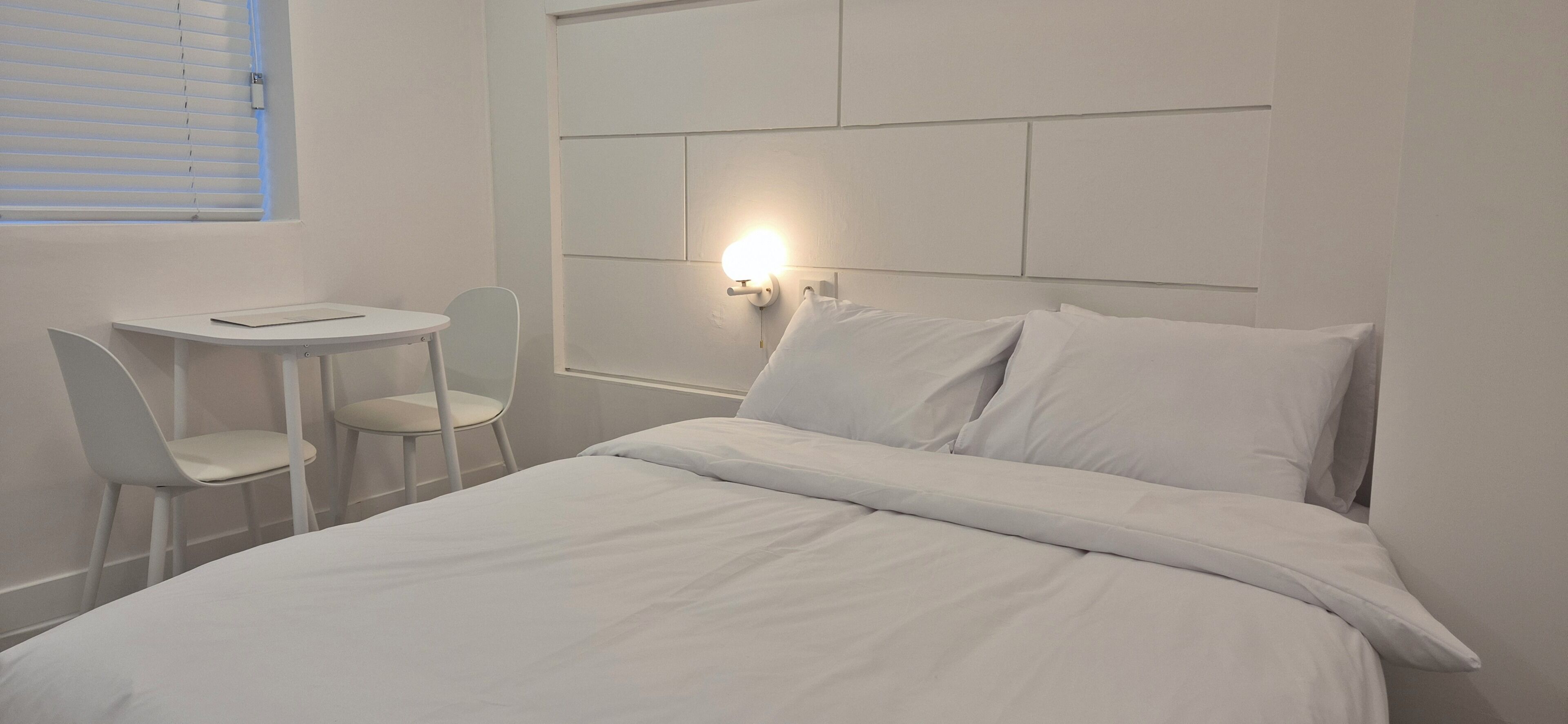 Quarto de hotel com cama, mesa de cabeceira, duas cadeiras e uma luminária na parede.