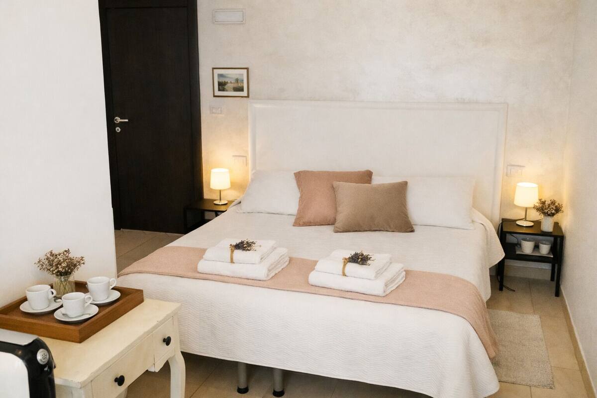 Uma cama bem arrumada com almofadas, uma mesa de cabeceira com abajur, e uma pequena mesa com xícaras e um vaso de flores.