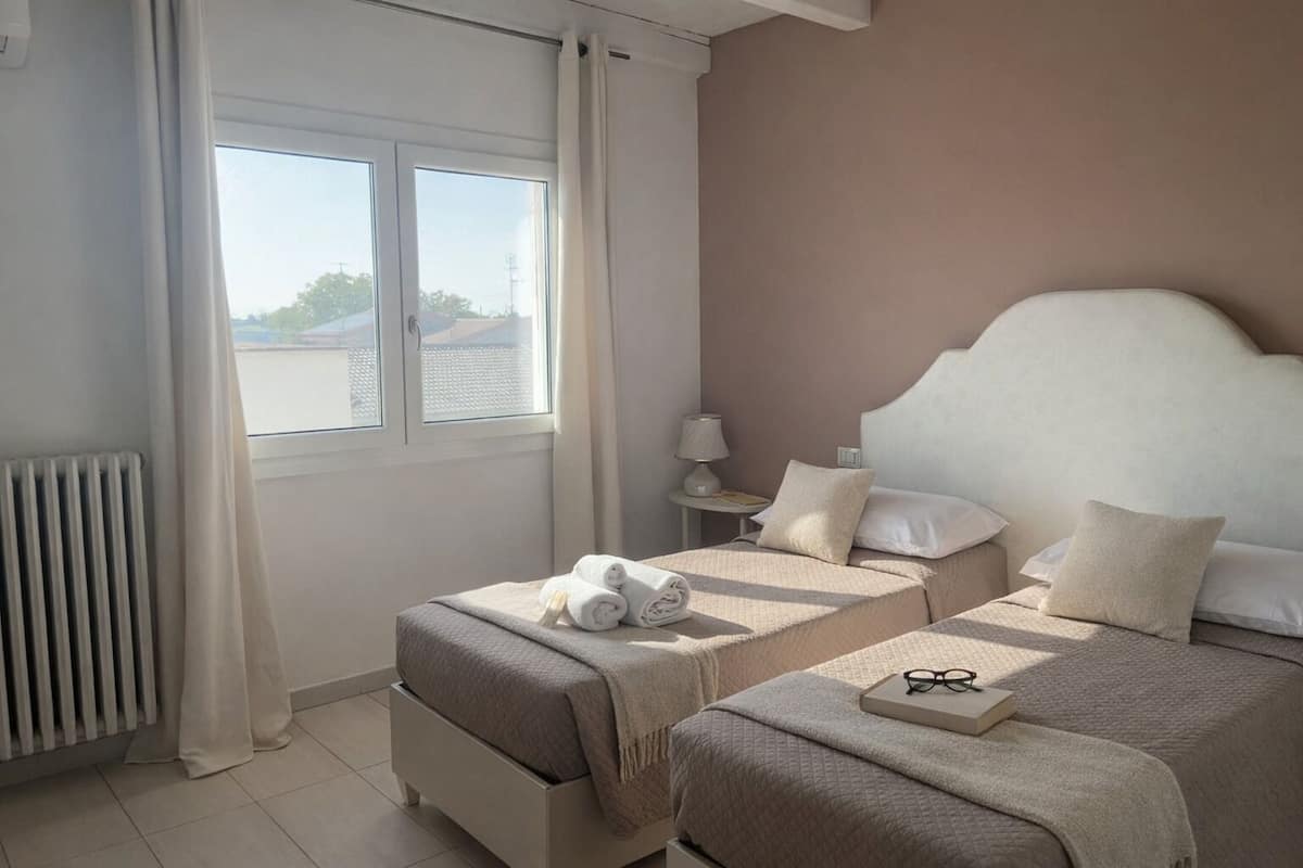 Quarto de hotel com duas camas, uma janela, um radiador e uma mesa de cabeceira com abajur.