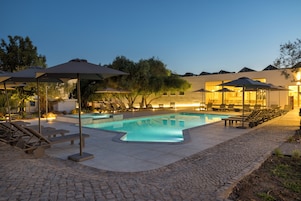 Piscina com espreguiçadeiras e guarda-sóis, rodeada por um pavimento e árvores.
