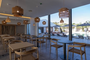 Restaurante com área externa e mesas de varanda com vista para uma piscina, com móveis de vime e luminárias pendentes.
