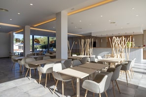 Um restaurante moderno com mesas e cadeiras dispostas num espaço amplo, janelas grandes com vista e um design limpo e contemporâneo.