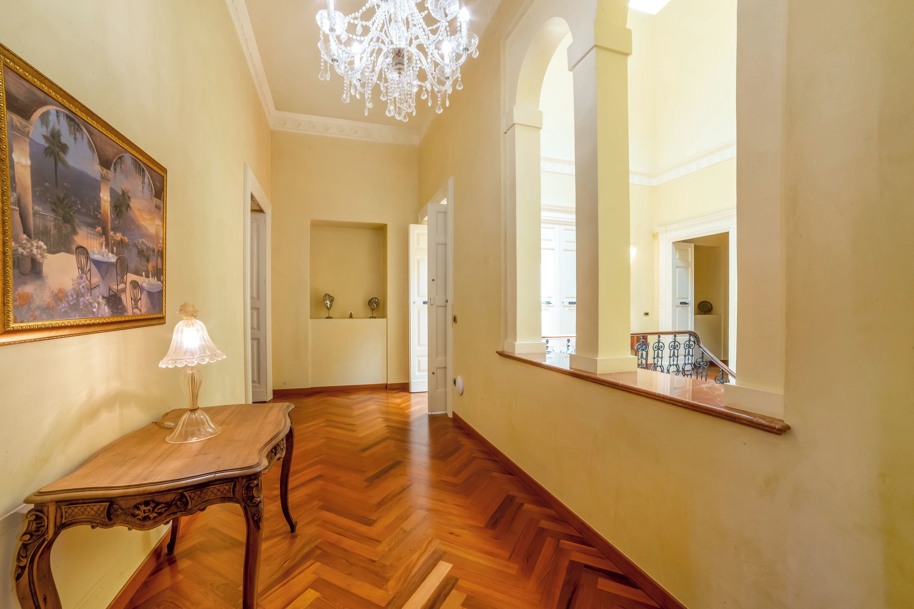 Um corredor com piso de madeira, um candelabro, um quadro e uma mesa com abajur.