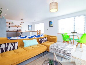Uma sala de estar moderna com um sofá amarelo, uma mesa de centro de vidro, cadeiras verdes e uma área de cozinha ao fundo.