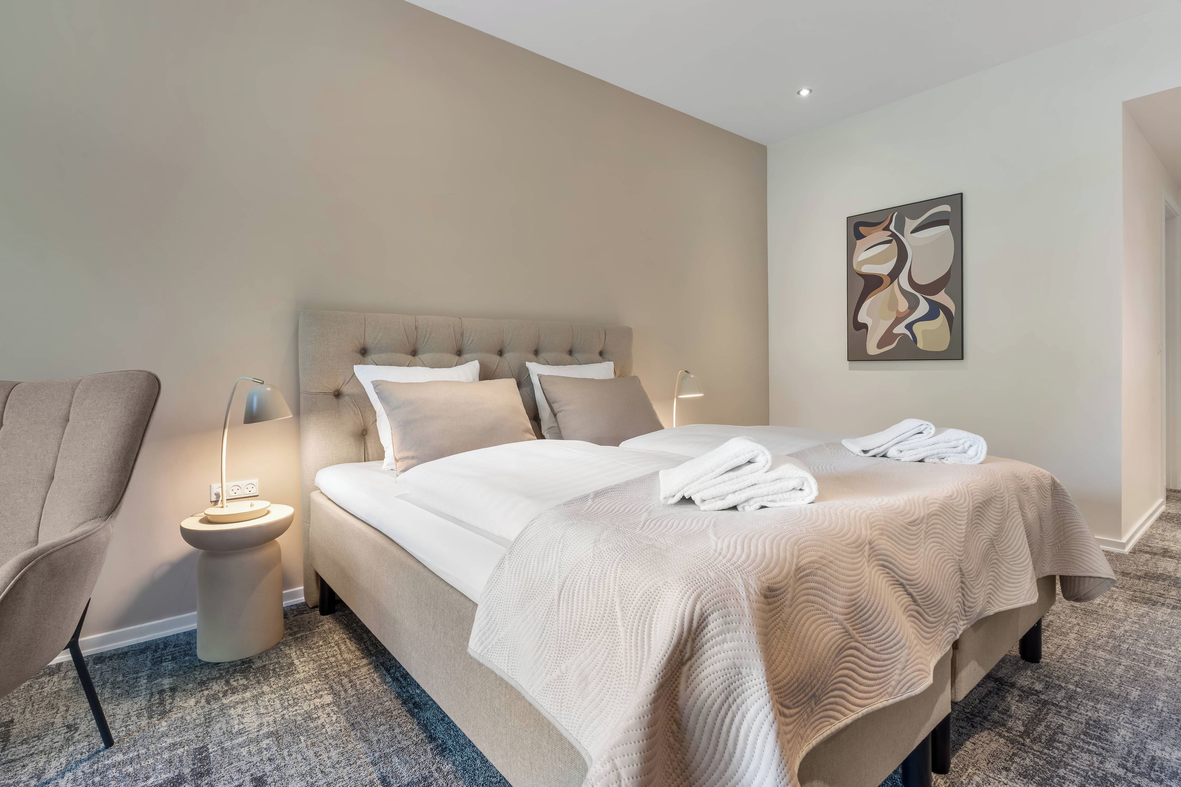 Quarto de hotel com cama, poltrona, mesa de cabeceira, abajur e um quadro na parede.