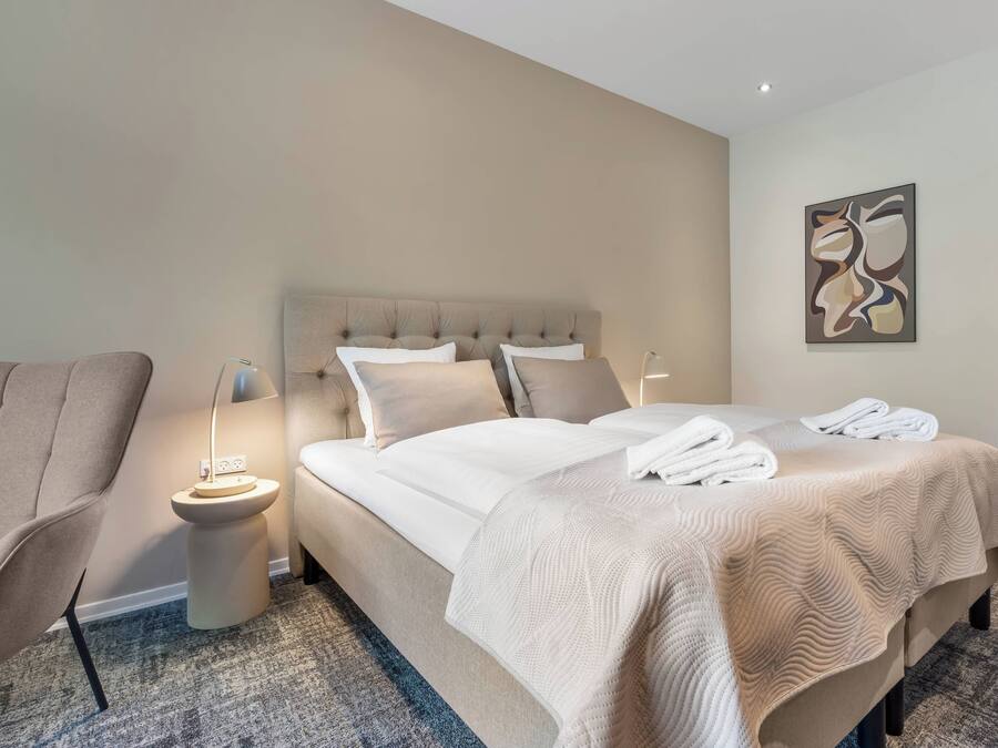 Quarto de hotel com cama, poltrona, mesa de cabeceira, abajur e um quadro na parede.