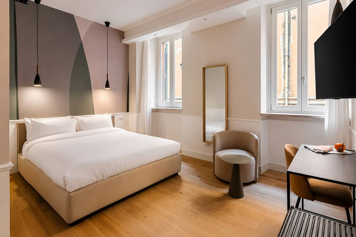 Quarto de hotel com cama, poltrona, mesa, espelho e luminárias pendentes.