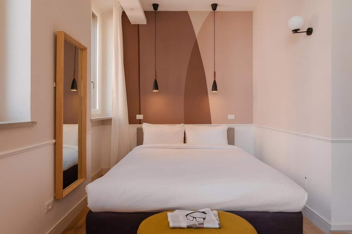 Quarto moderno com uma cama grande, luminárias de cabeceira, um espelho e um pequeno banquinho com óculos em cima.