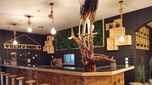 Uma área de bar com uma escultura central de árvore em madeira, uma placa com a inscrição 'BEM VINDO' e vários bancos.