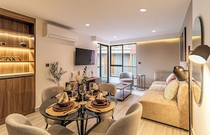 Uma sala de estar moderna com um sofá, uma mesa de centro, uma mesa de jantar posta para quatro pessoas e uma televisão na parede.