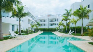 Um hotel moderno com piscina central, rodeado por palmeiras e edifícios brancos.