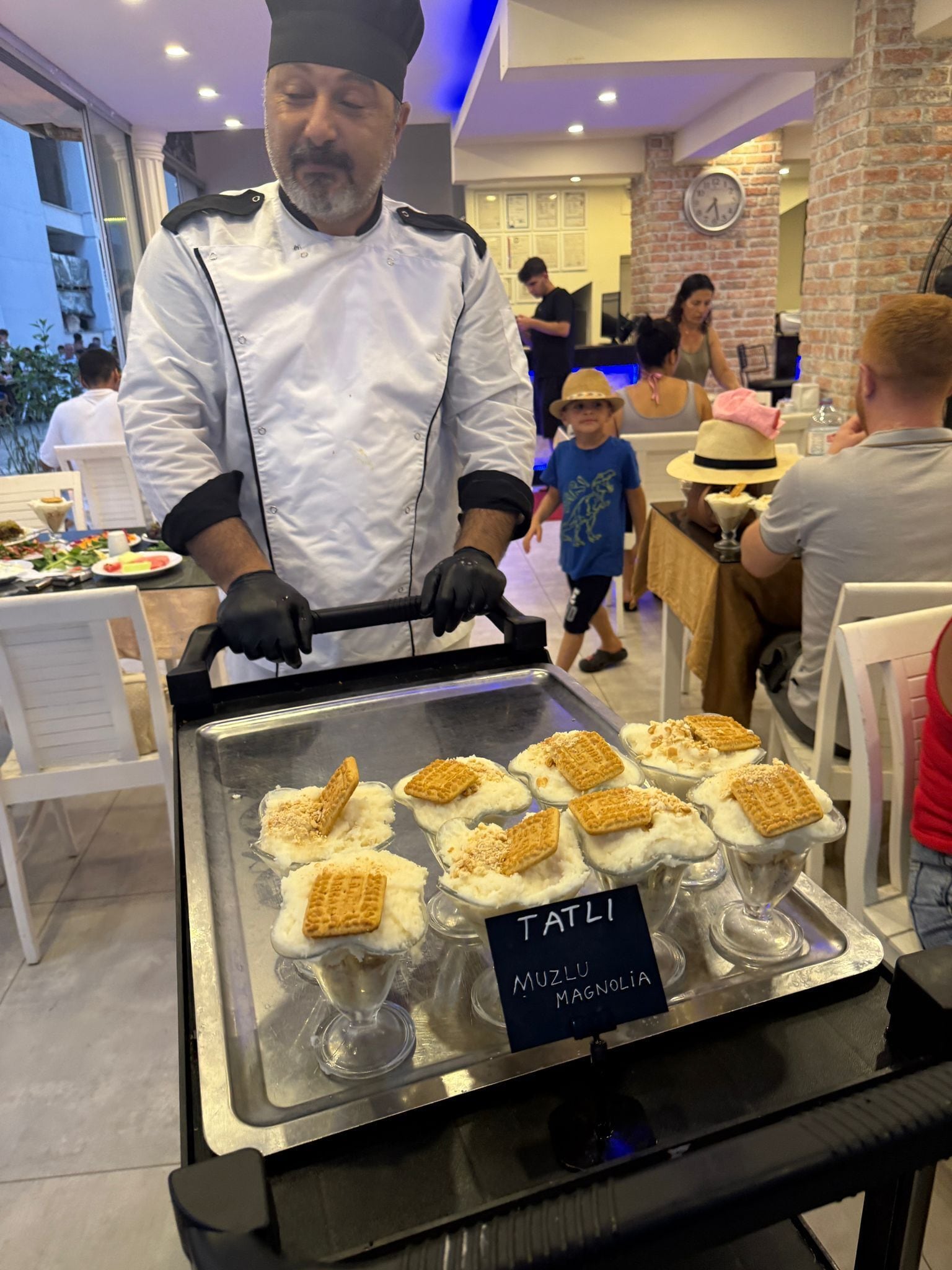 Um chef servindo uma sobremesa com bolachas em um prato com a etiqueta 'TATLI MUZLU MAGNOLIA'.