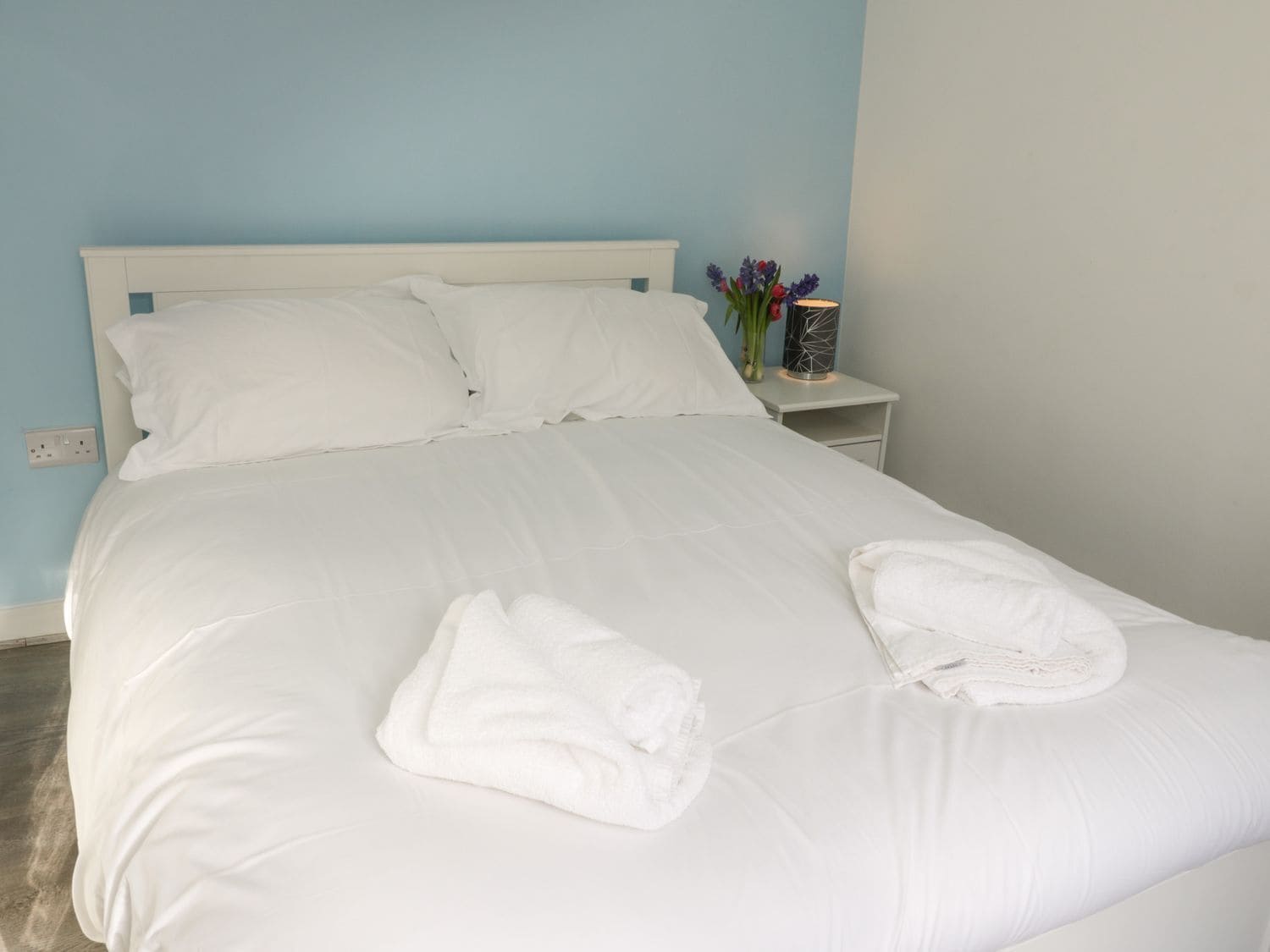 Uma cama bem arrumada com lençóis e toalhas brancas, uma mesa de cabeceira com um vaso de flores e uma tomada na parede.