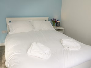 Uma cama bem arrumada com lençóis e toalhas brancas, uma mesa de cabeceira com um vaso de flores e uma tomada na parede.
