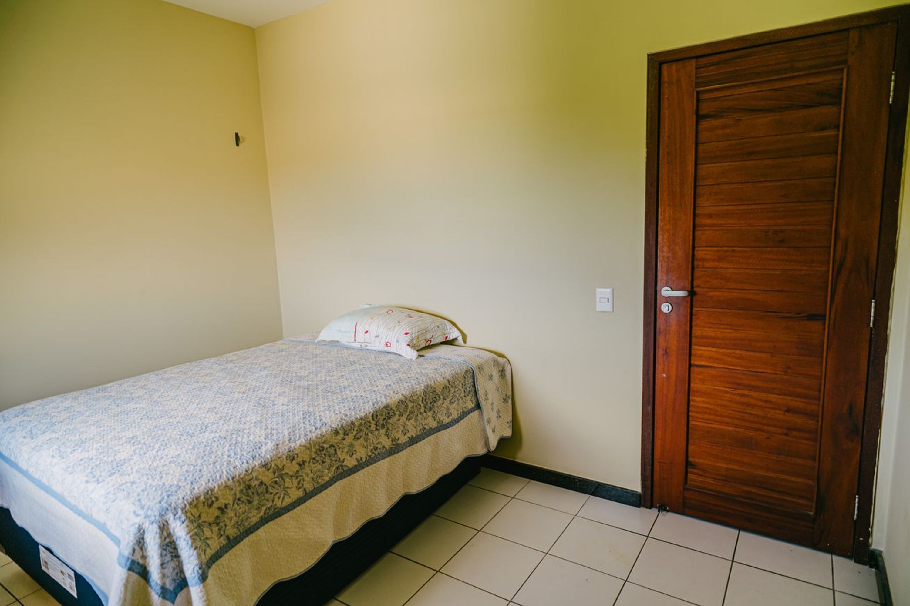 Quarto com uma cama, porta de madeira e chão de azulejos.