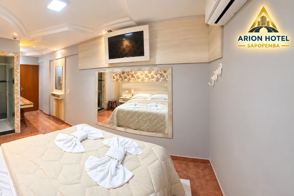 Quarto de hotel com cama, televisão, espelho e chuveiro.