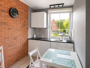 Uma cozinha compacta com parede de tijolos, um relógio, uma janela com vista para edifícios e uma mesa com toalha de mesa estampada.