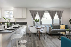 Cozinha moderna com armários brancos, sala de jantar com sofá cinza e espaço de bar com banquetas brancas.