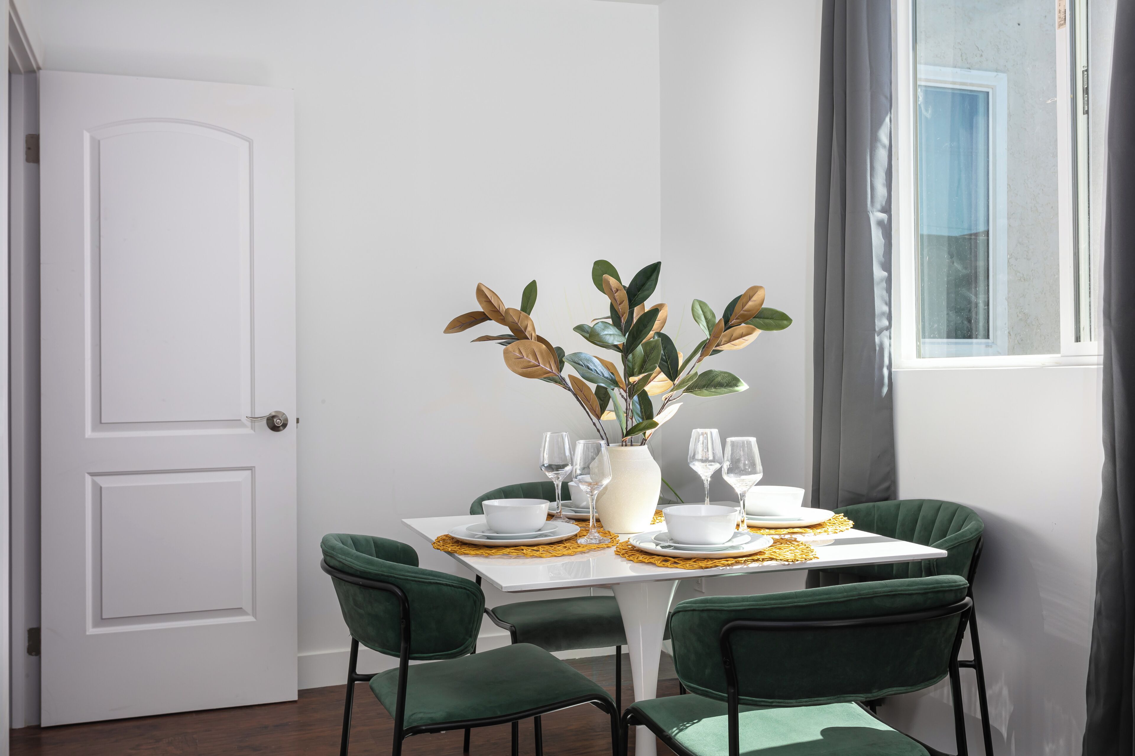 Uma área de refeições com uma mesa branca, cadeiras verdes, um vaso com folhas e uma janela com cortinas.