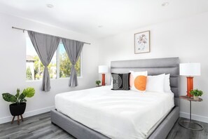 Um quarto moderno com uma cama grande, cortinas cinzas, uma planta em vaso e um quadro na parede.