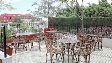 Mobiliário de exterior com cadeiras e mesas de metal decorativas, chão em mosaicos e vista para edifícios e árvores.