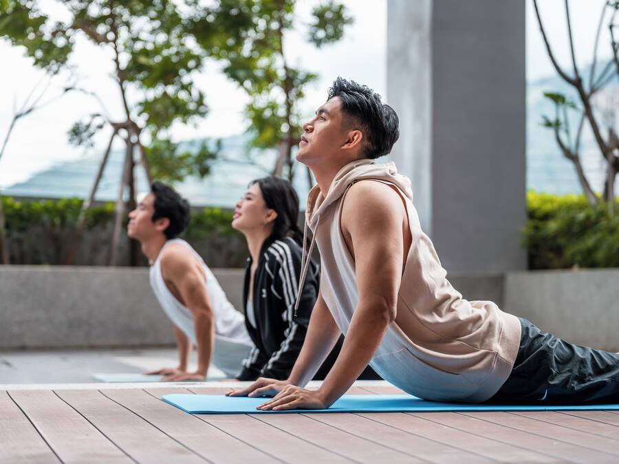 Três pessoas praticando yoga em um deck de madeira, com vista para árvores e um edifício ao fundo.