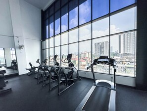 Uma academia com esteiras, elípticos e um ventilador, com vista para a cidade através de grandes janelas.