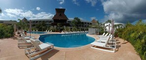Uma piscina com espreguiçadeiras, um edifício com telhado em forma de pirâmide e palmeiras.