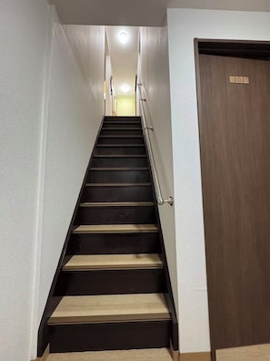 Uma escada com corrimão de madeira, uma porta com a inscrição '102' e uma luz no topo.