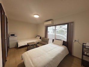 Quarto pequeno e bem iluminado com duas camas, uma escrivaninha, uma cadeira, micro-ondas e janela com persianas.