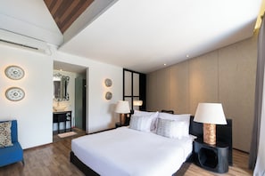 Quarto de hotel moderno com uma cama grande, luminárias de cabeceira, um pequeno espaço de estar e um banheiro visível por uma porta aberta.