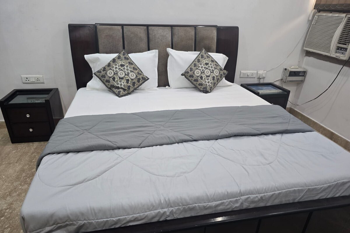 Cama bem arrumada com colcha cinza e duas almofadas decorativas, mesa de cabeceira com abajur e ar condicionado fixado na parede.