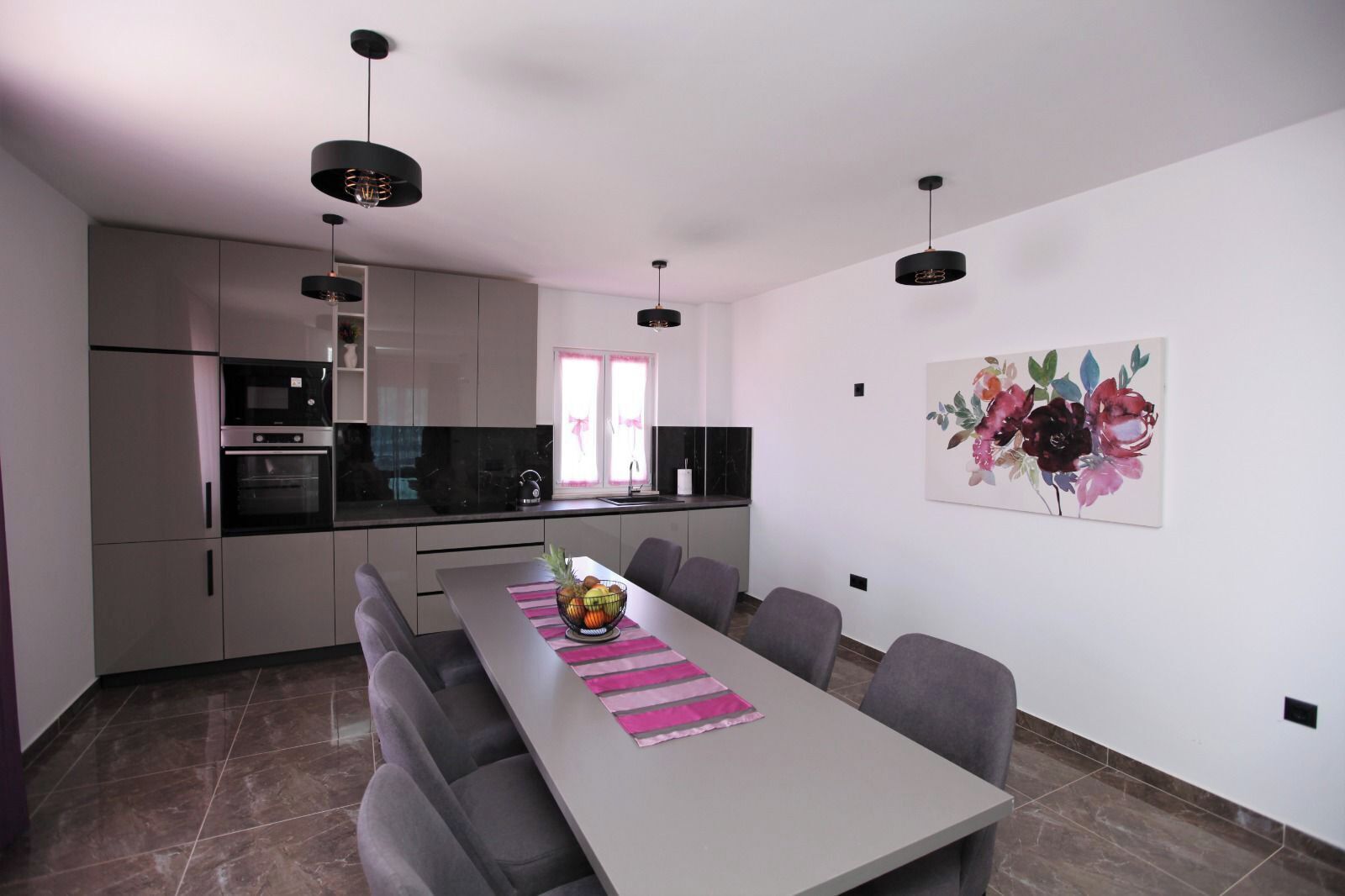 Uma cozinha moderna com eletrodomésticos de aço inoxidável, uma área de jantar com uma mesa comprida e cadeiras, e um quadro floral na parede.