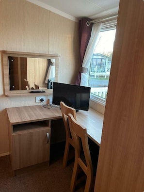Quarto compacto com escrivaninha de madeira, cadeira, espelho e janela com cortinas.