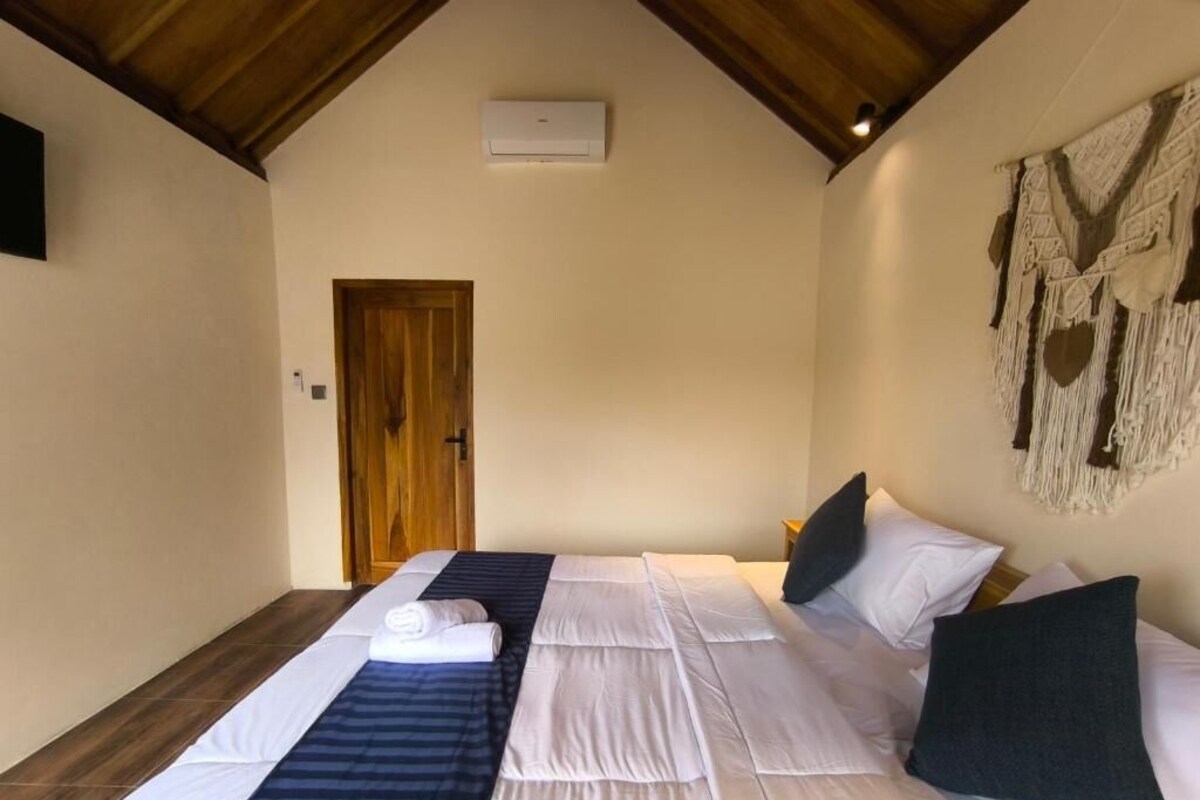 Quarto com teto de madeira, cama com roupa de cama branca e listrada, porta de madeira e decoração na parede.