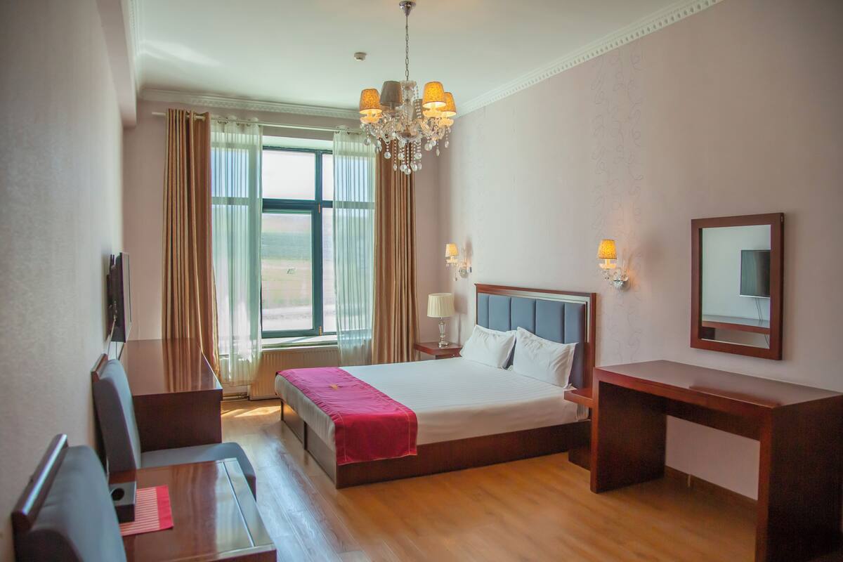Quarto de hotel com cama, escrivaninha, cadeira e uma grande janela com cortinas.
