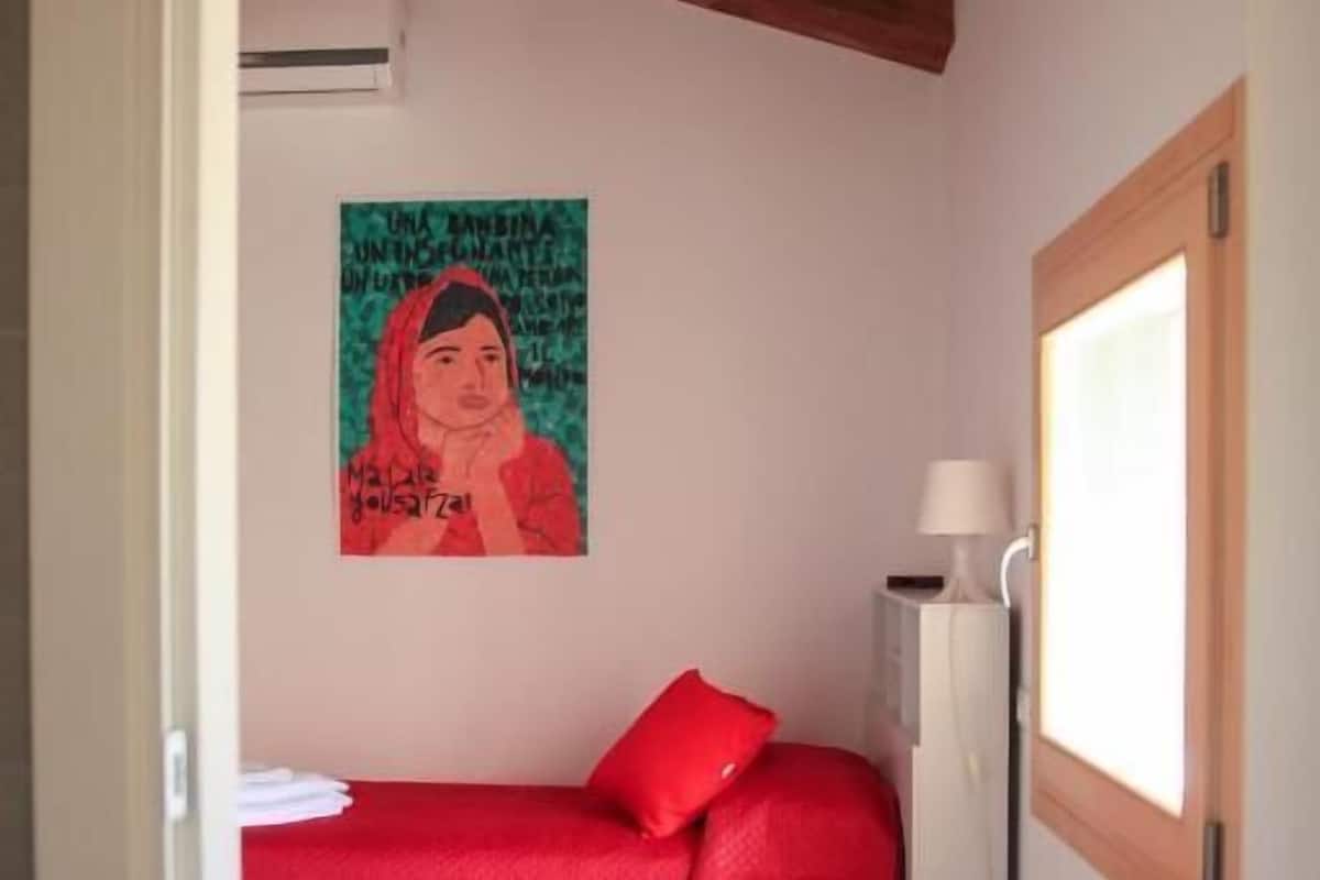 Um quarto pequeno com cama vermelha, tapete azul, luminária branca e um pôster na parede.
