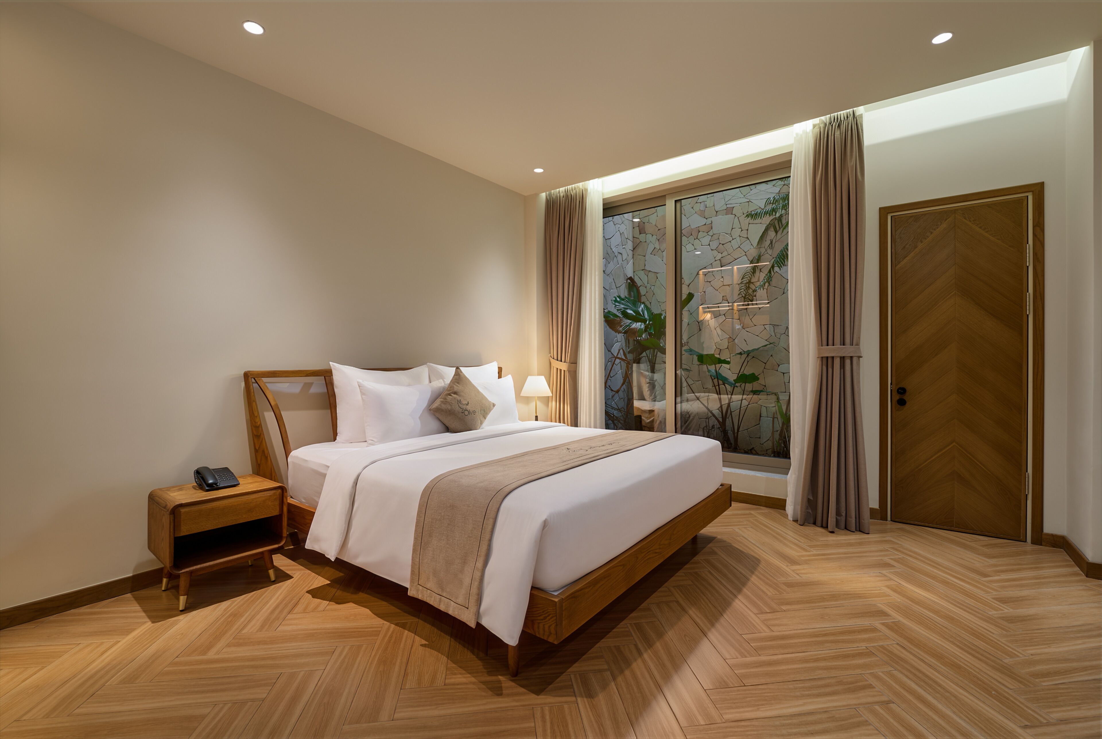 Quarto de hotel com uma cama grande, uma mesa de cabeceira com telefone, uma janela com cortinas e uma porta de madeira.